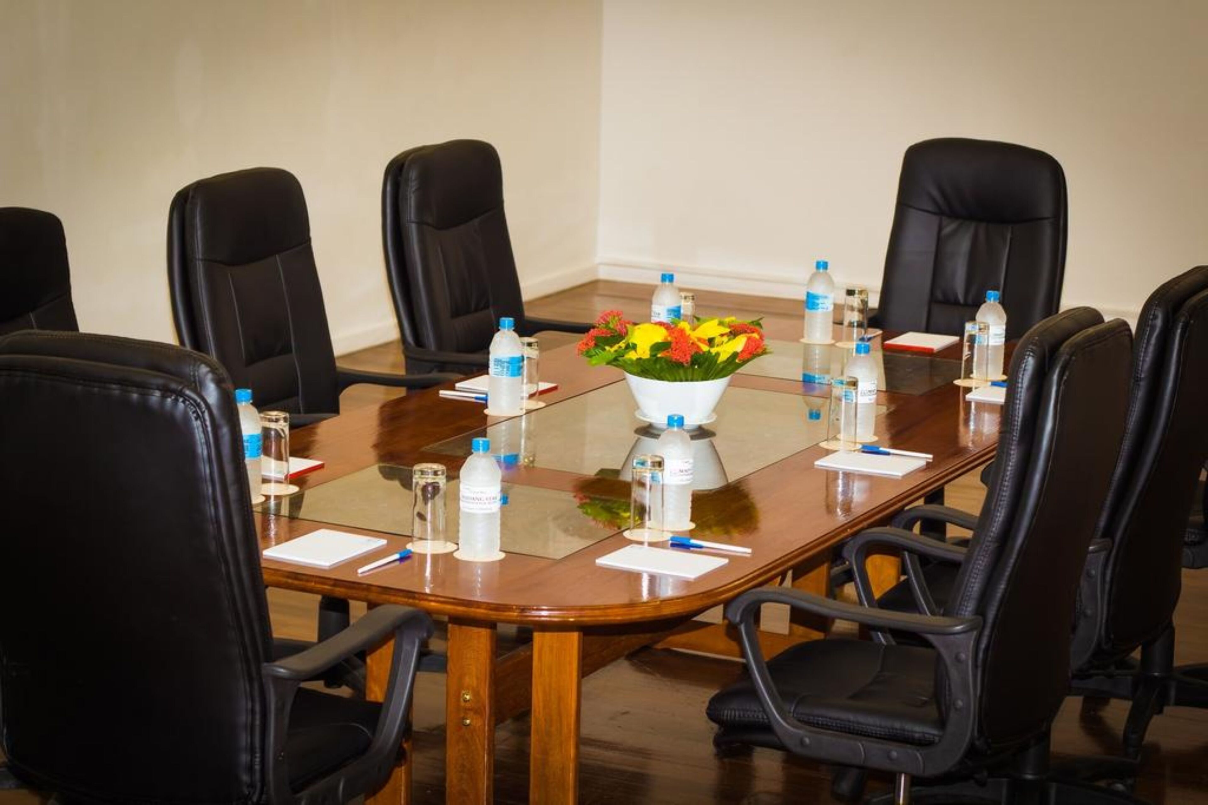 Uma sala de conferência com mesa de vidro, cadeiras de couro preto, garrafas de água e um arranjo de flores.