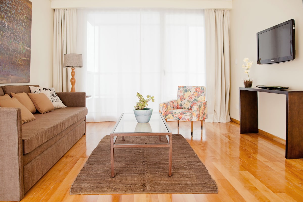 Uma sala de estar com sofá, mesa de centro, poltrona, televisão e uma janela grande com cortinas.