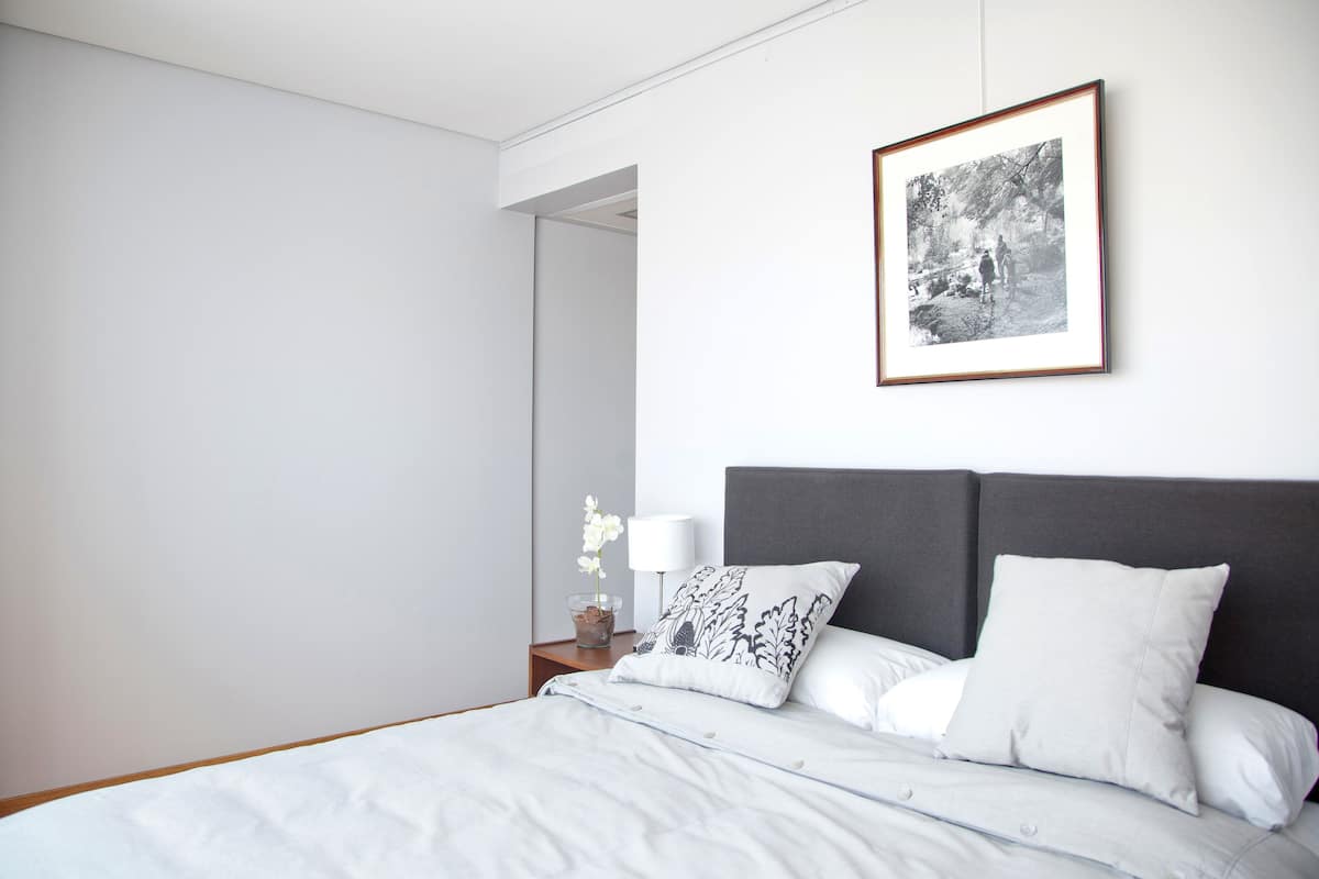 Uma cama bem arrumada com lençóis brancos e duas almofadas, uma mesa de cabeceira com abajur e um vaso de flores, e um quadro na parede.