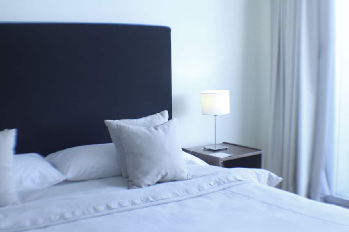 Uma cama bem arrumada com lençóis e almofadas brancas, uma mesa de cabeceira com abajur e uma janela com cortinas.