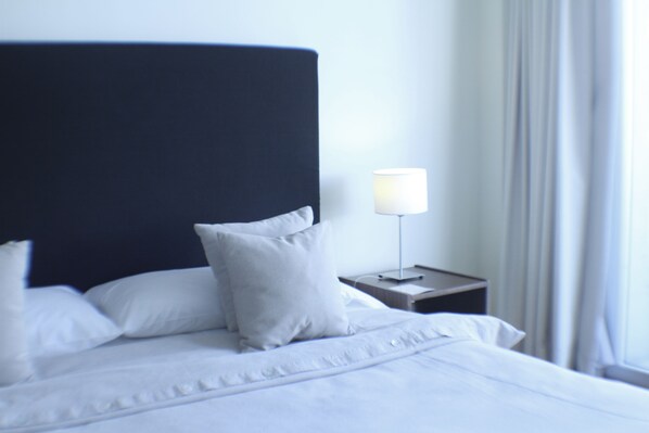 Uma cama bem arrumada com lençóis e almofadas brancas, uma mesa de cabeceira com abajur e uma janela com cortinas.