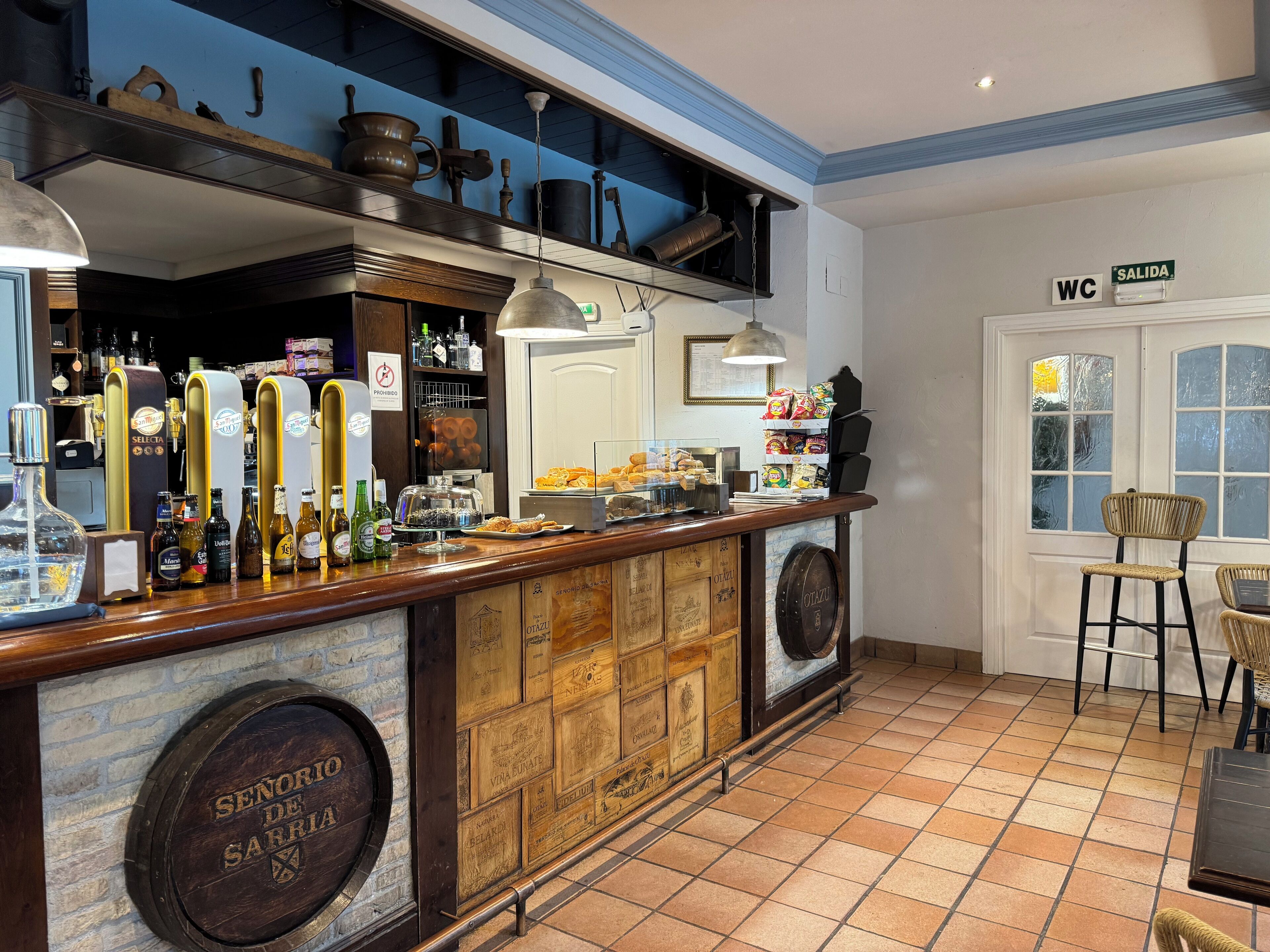 Uma área de bar com balcão de madeira, prateleiras com garrafas e um aviso indicando um banheiro.