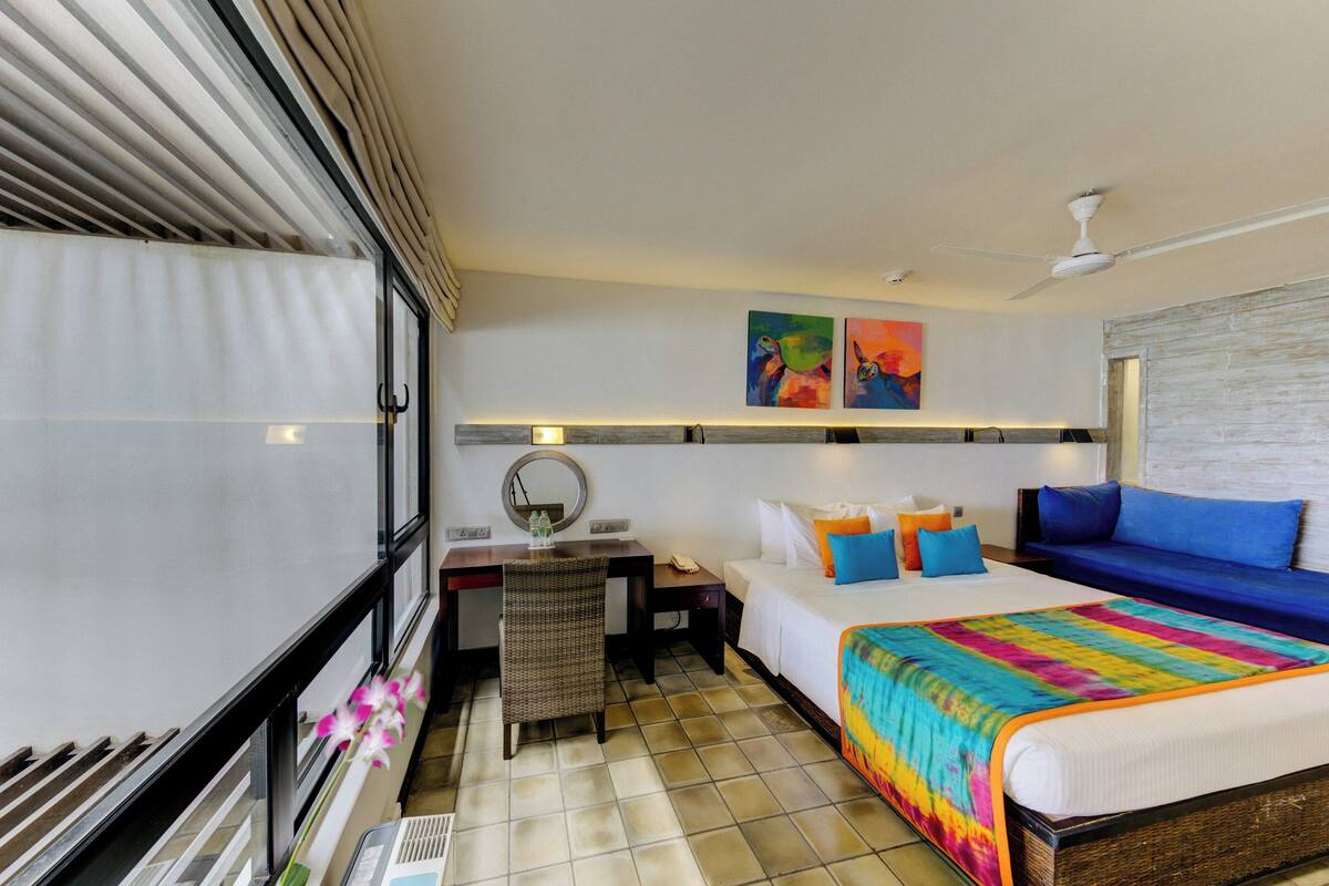 Quarto de hotel com cama, escrivaninha, cadeira, ventoinha no teto e obras de arte na parede.
