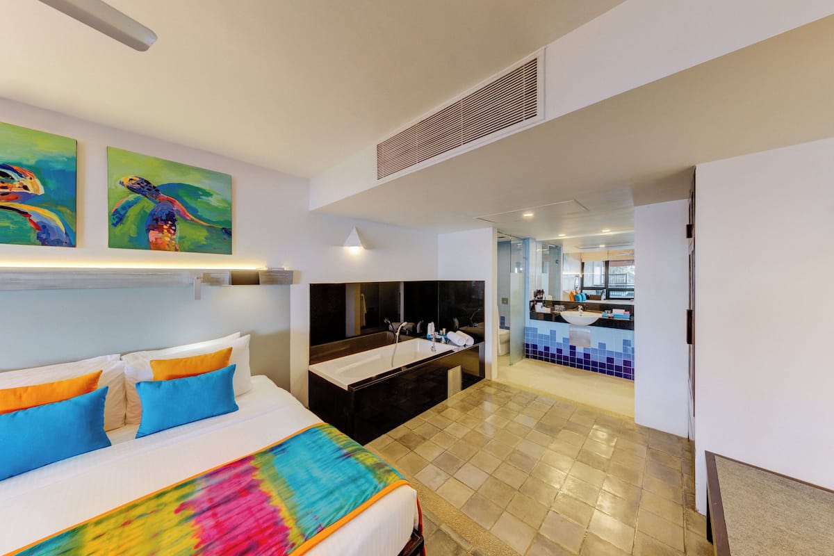 Quarto de hotel com uma cama grande, banheiro com pia e espelho, e vista para o quarto vizinho.