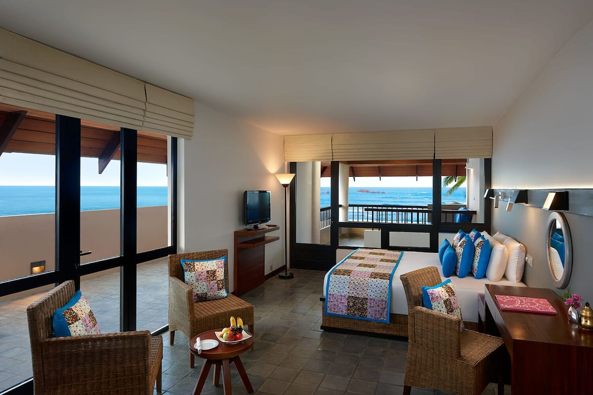 Varanda ampla com vista para o oceano, mobiliada com cadeiras de vime e uma pequena mesa com frutas.