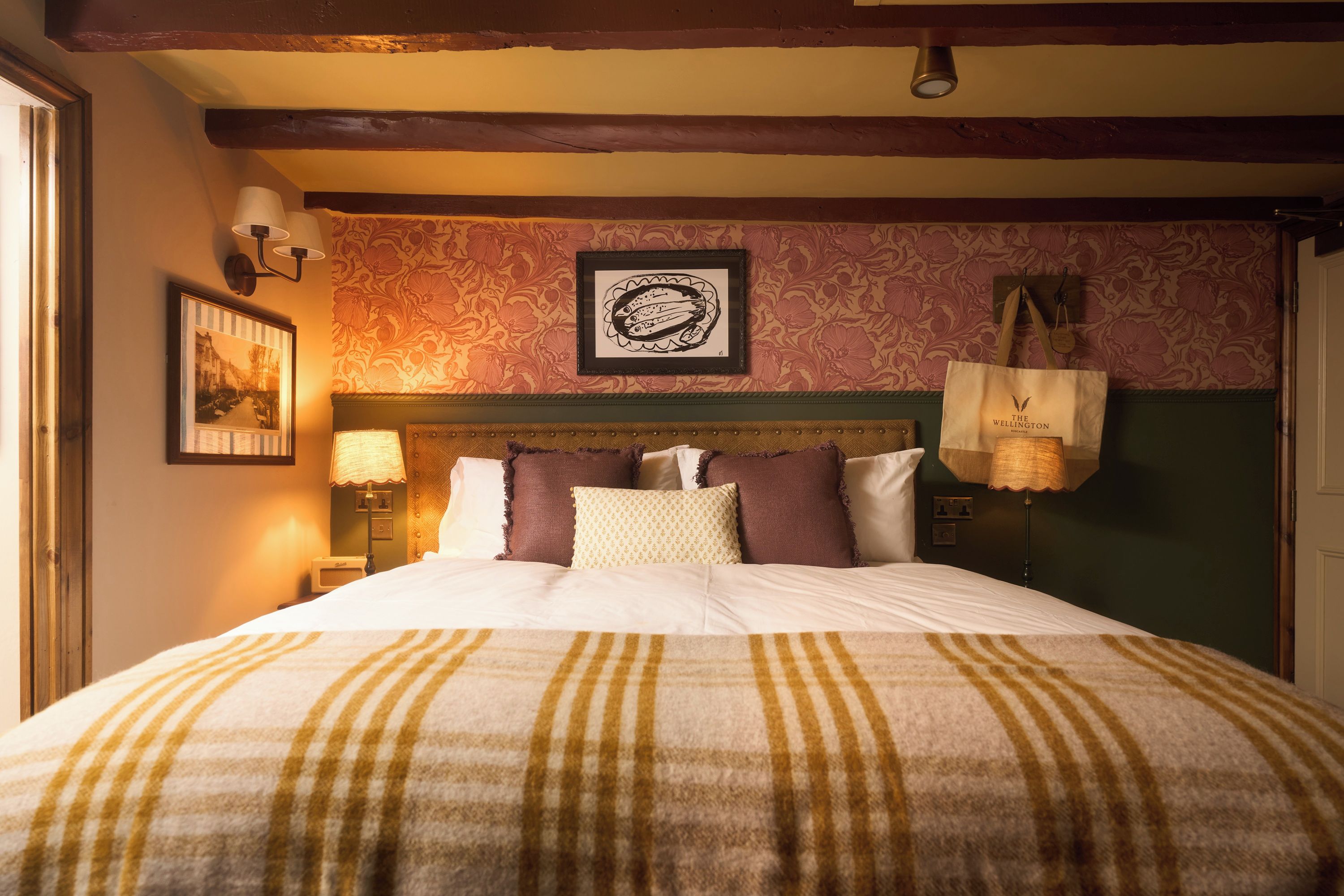 Quarto de hotel com uma cama grande, cabeceira, quadro emoldurado, abajur e parede com papel de parede estampado.