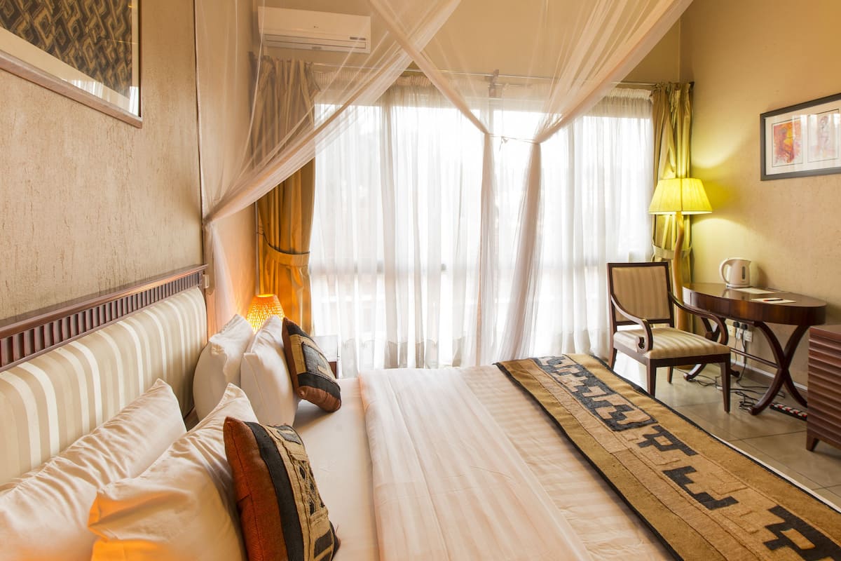 Quarto de hotel com cama, escrivaninha, cadeira, luminária e janela com cortinas leves.