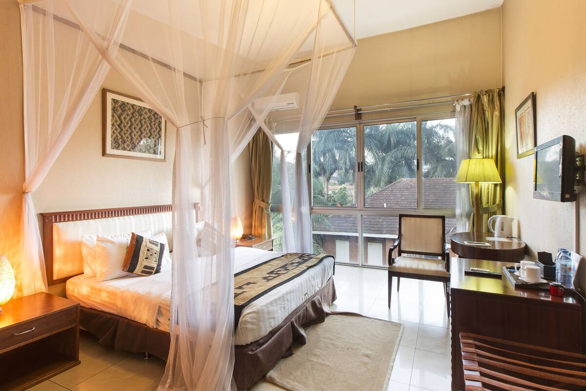 Quarto com cama, mesa de cabeceira, abajur e vista para palmeiras.
