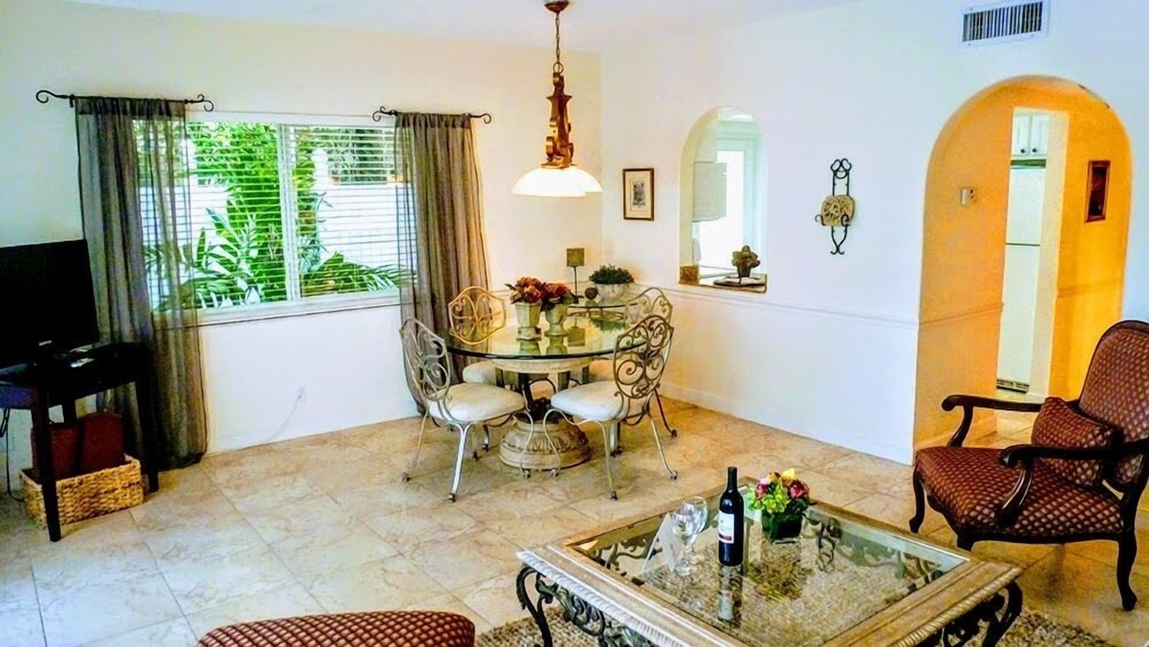 Uma sala de estar com uma mesa de centro de vidro, uma mesa de jantar com cadeiras, uma televisão e uma grande janela com cortinas.