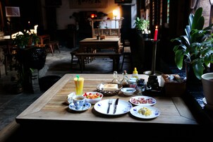 Mesa de jantar posta com pratos, talheres e uma vela, rodeada por cadeiras e plantas em vasos num ambiente com pouca luz.