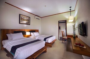 Quarto de hotel com duas camas, cabeceira de madeira, mesa de cabeceira com abajur, quadro pendurado na parede e área de jantar com mesa e cadeiras.