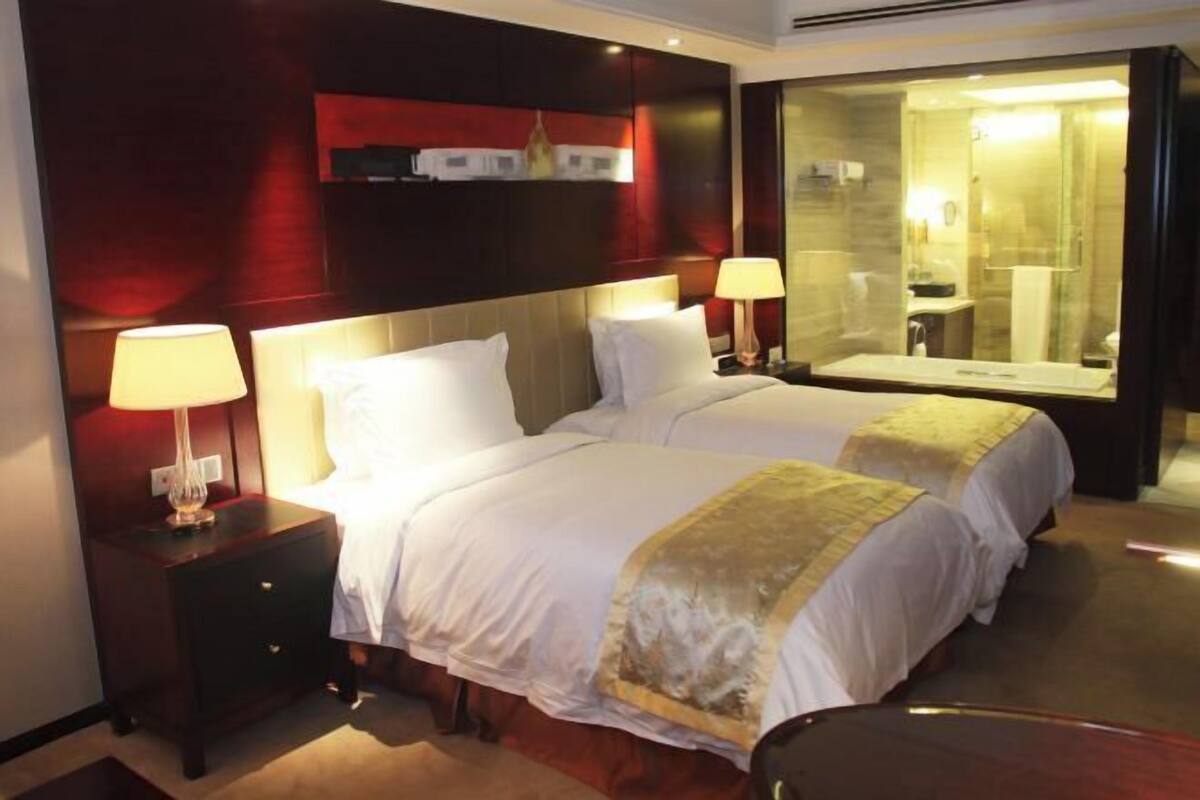 Quarto de hotel com duas camas, uma mesa de cabeceira com luminária, um espelho grande e um banheiro visível através de uma divisória de vidro.