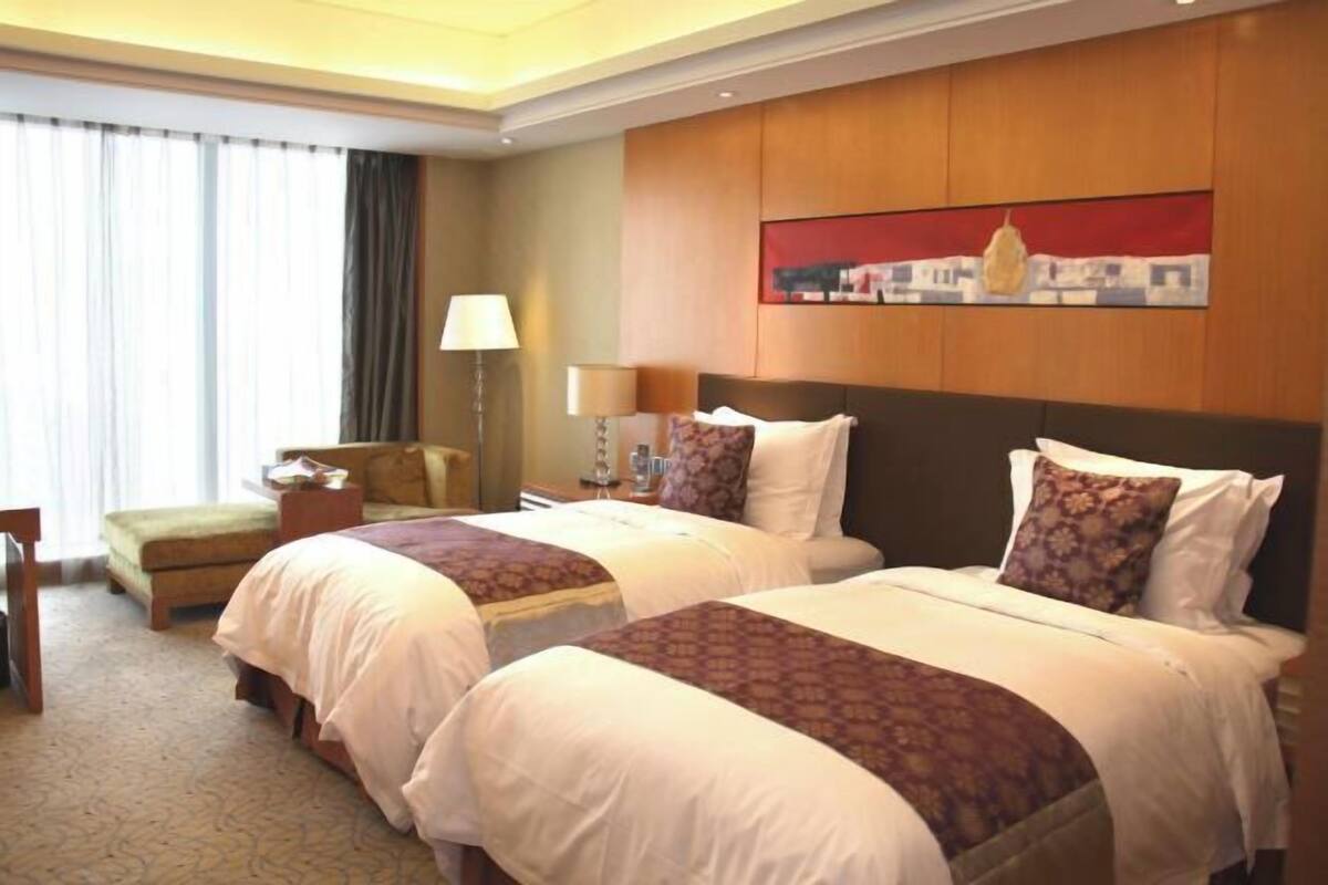 Quarto de hotel com duas camas, uma escrivaninha, uma cadeira, um abajur e um quadro na parede.