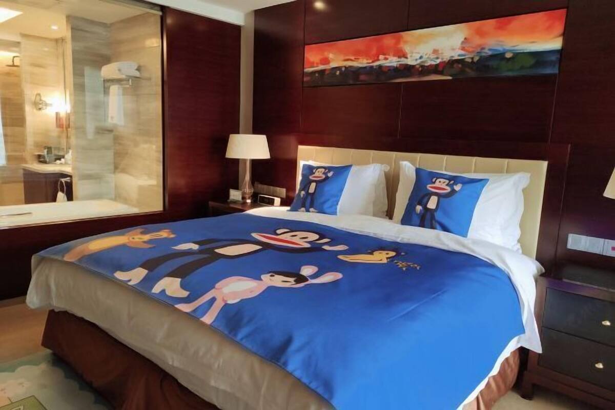 Quarto de hotel com uma cama grande, mesa de cabeceira, luminária, pintura na parede e banheiro visível por uma porta de vidro.