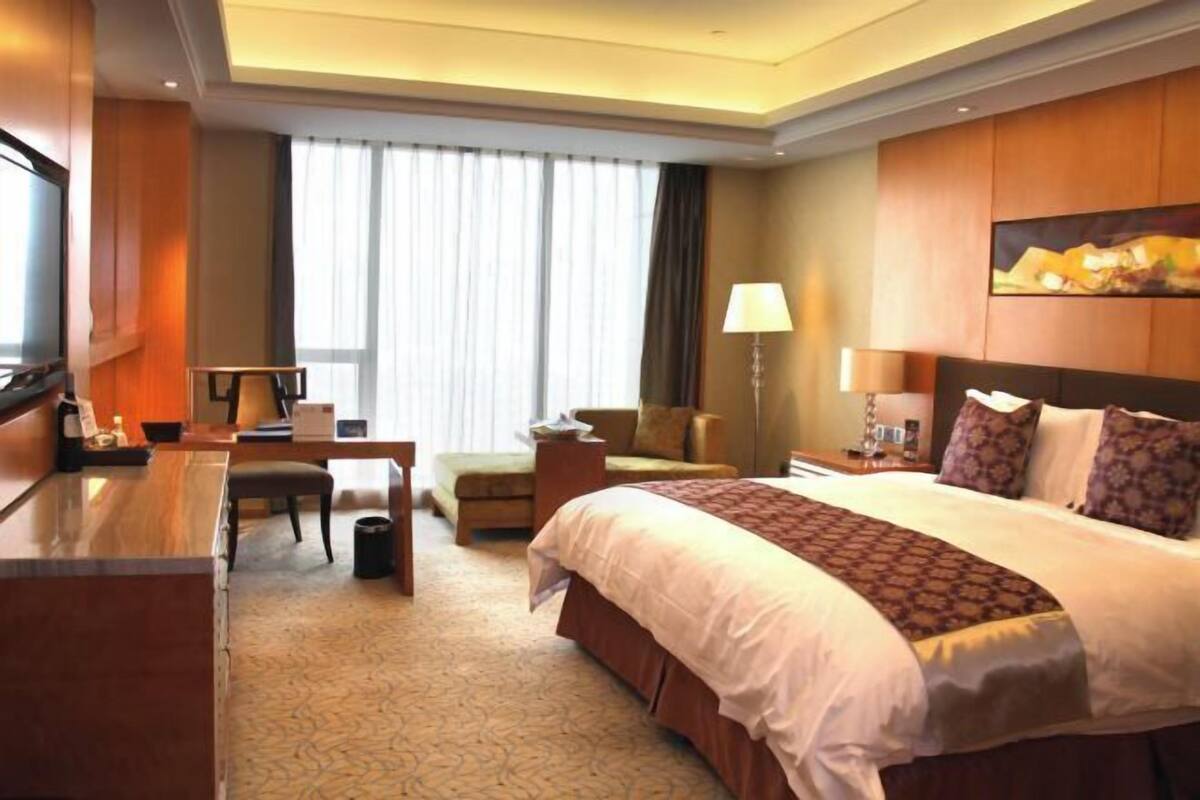 Quarto de hotel com cama, escrivaninha, cadeira, abajur e uma janela grande com cortinas.