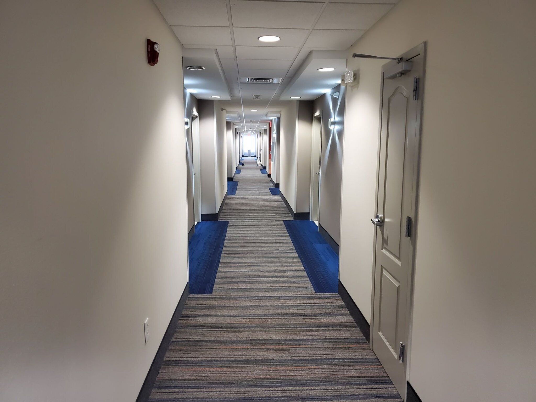 Corredor de hotel com piso coberto por carpete, portas em ambos os lados e iluminação no teto.