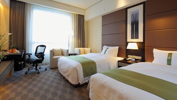 Quarto de hotel com duas camas, uma secretária com cadeira, uma luminária e uma janela com cortinas.