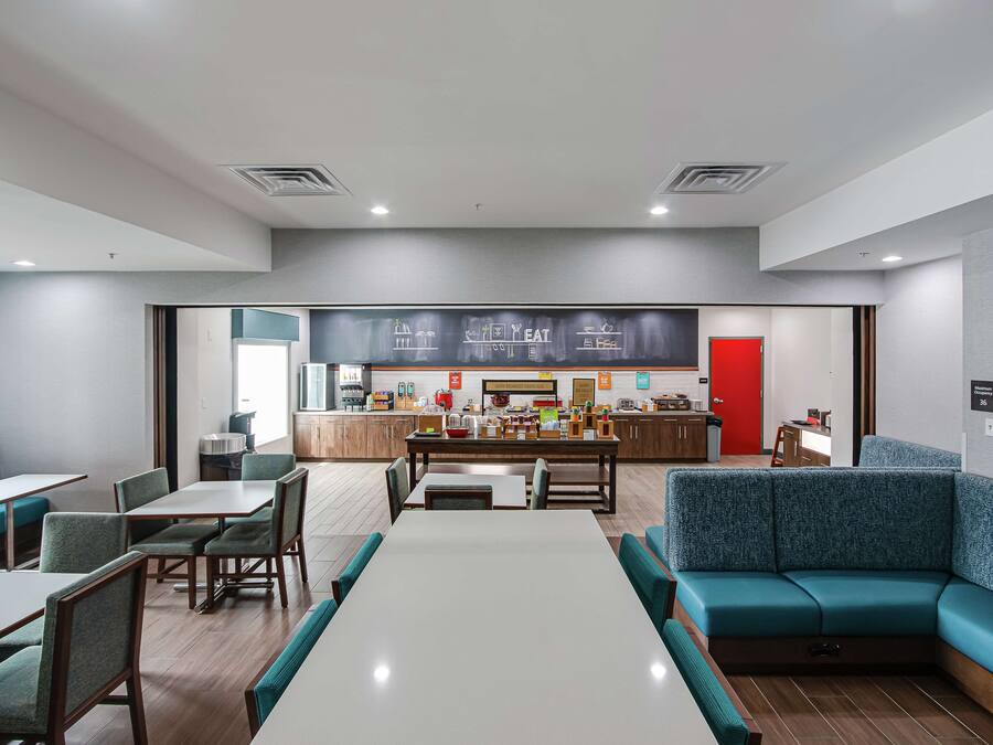 Uma área de refeições moderna com mesas e cadeiras, um balcão com produtos alimentares e uma porta vermelha ao fundo.