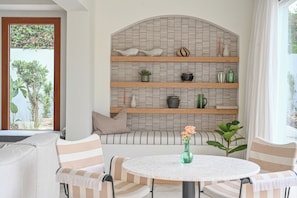 Quarto com mesa redonda, sofá riscado e prateleira na parede com objetos decorativos.