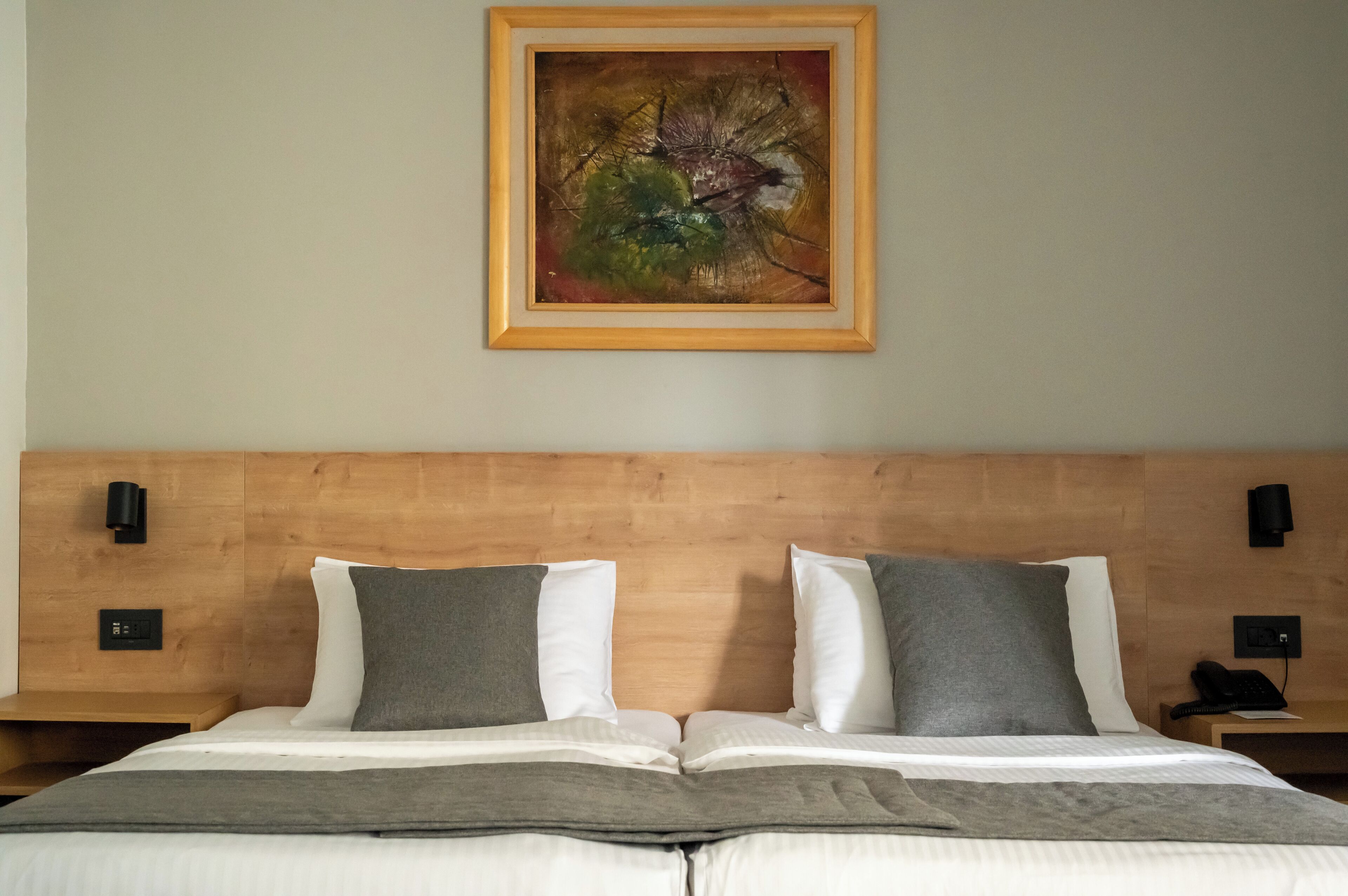 Uma cama de casal com roupa de cama branca e cinza, duas mesas de cabeceira com abajures pretos, uma cabeceira de madeira e uma pintura abstrata emoldurada acima da cama.