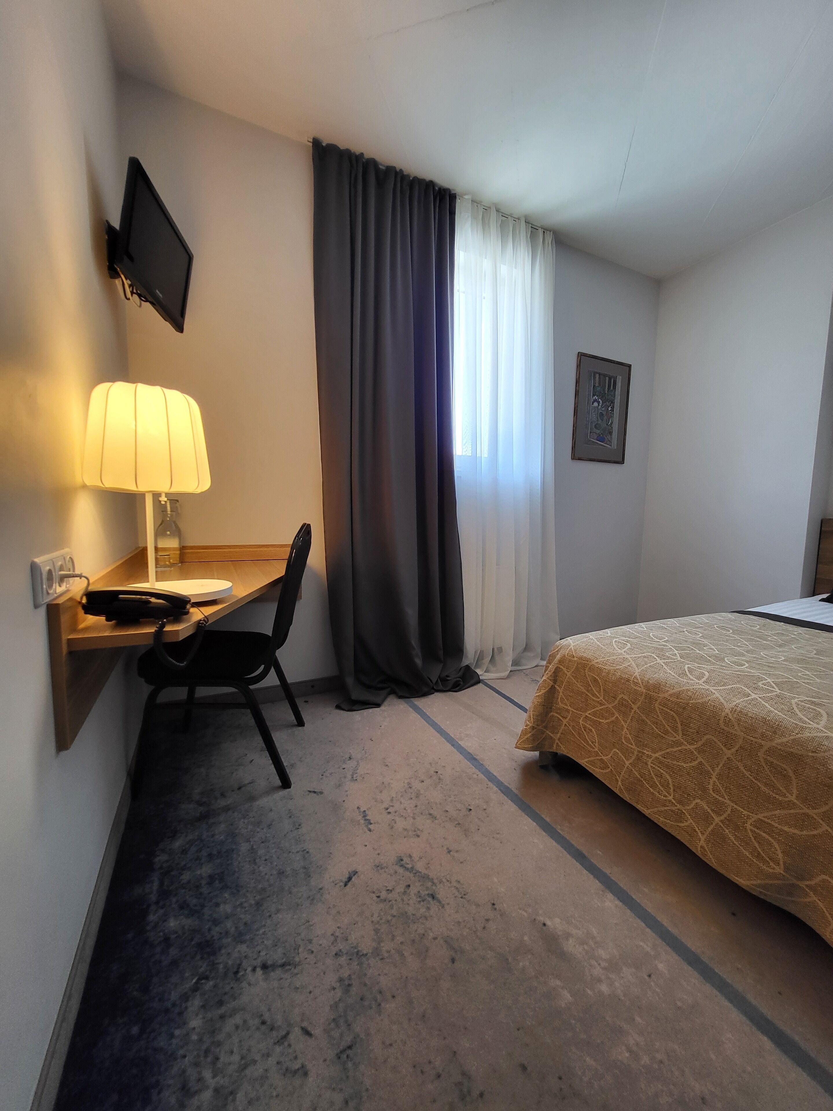 Quarto de hotel com escrivaninha, cadeira, cama, TV e cortinas.