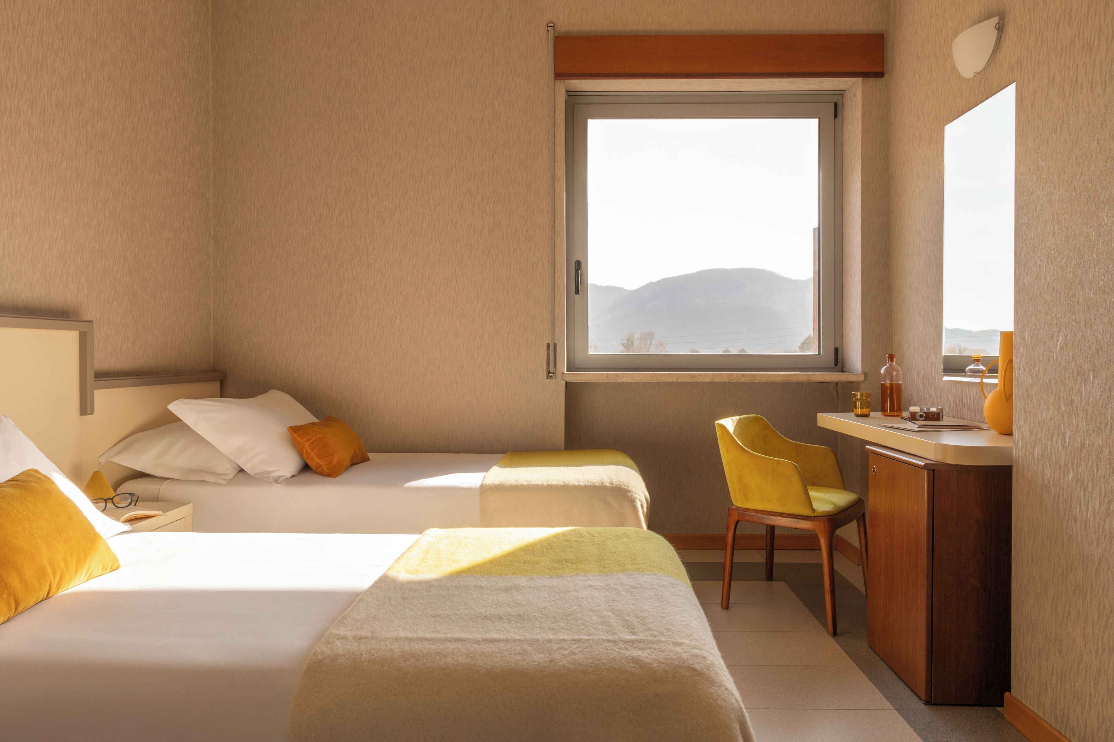 Quarto de hotel com duas camas, uma escrivaninha, uma cadeira, janela com vista e um espelho.