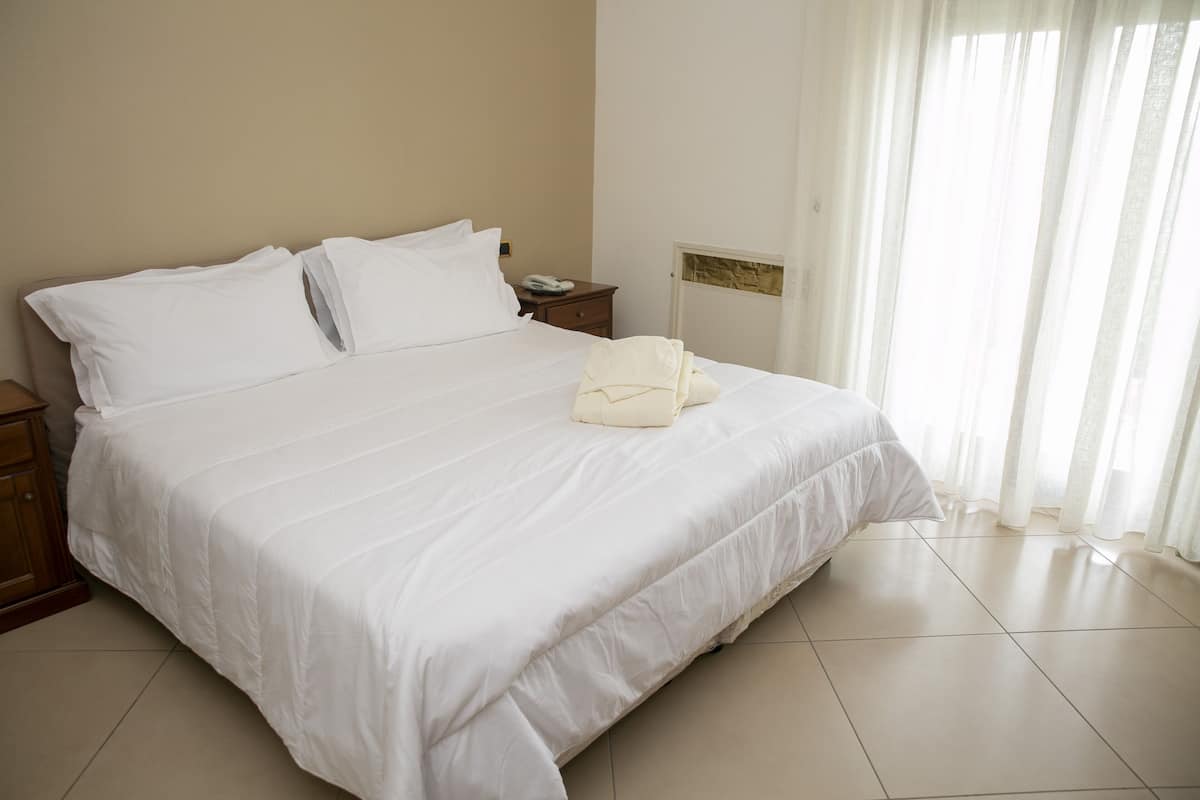 Uma cama bem arrumada com lençóis brancos, uma mesa de cabeceira com telefone e uma janela com cortinas leves.