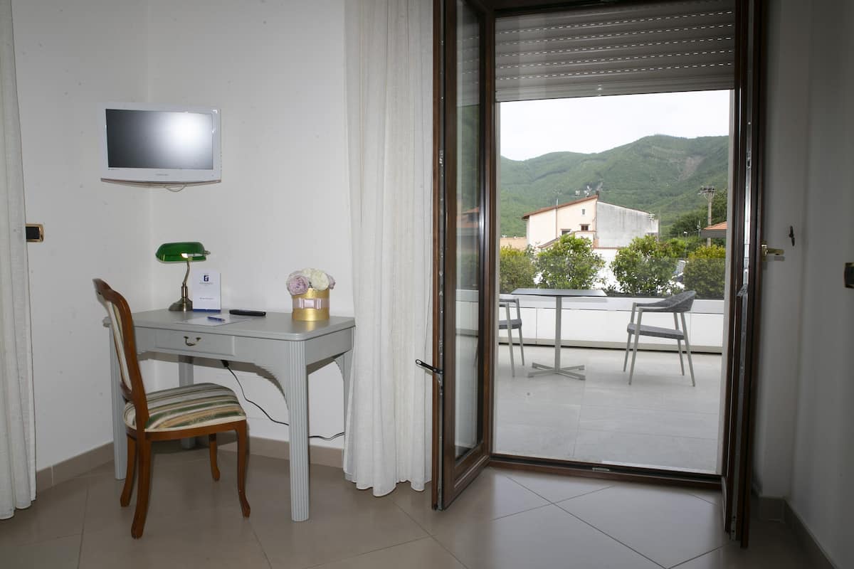 Quarto com escrivaninha, cadeira, televisão e porta de vidro deslizante para uma varanda ao ar livre com mesa e cadeiras.