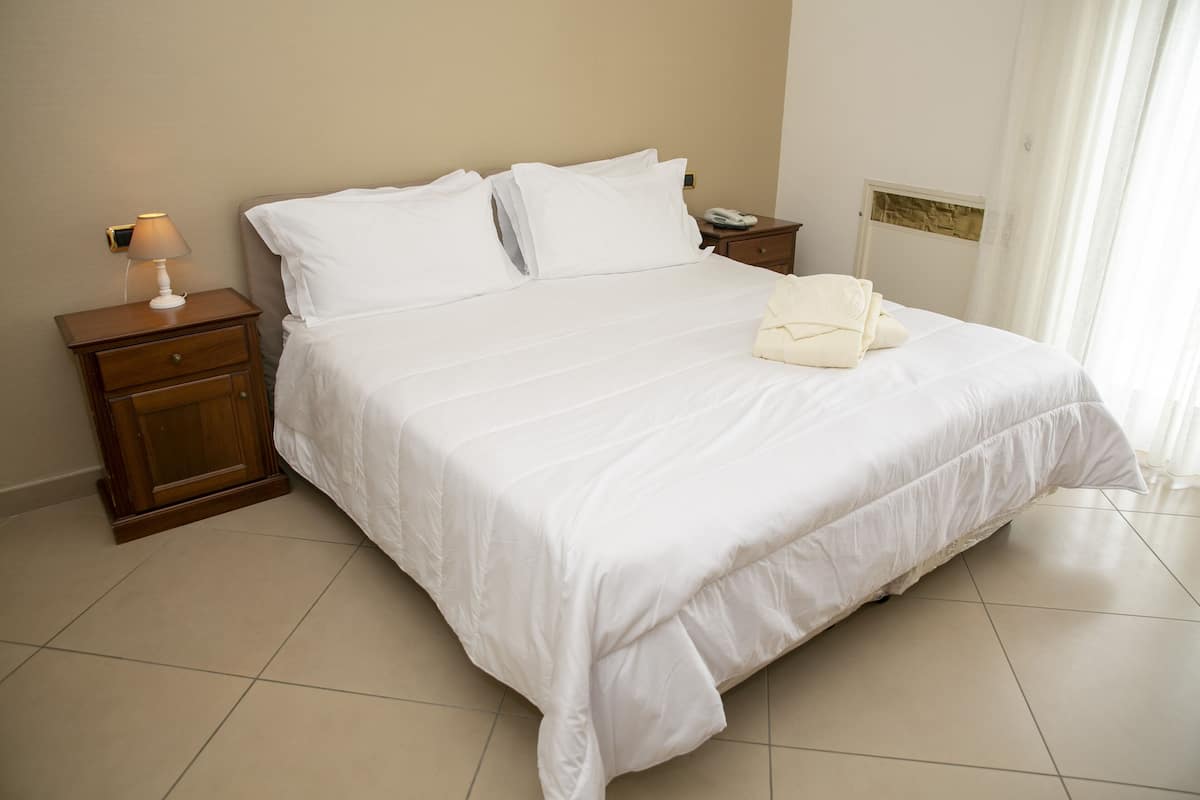 Cama bem arrumada com lençóis brancos, mesa de cabeceira com abajur e toalha dobrada sobre a cama.
