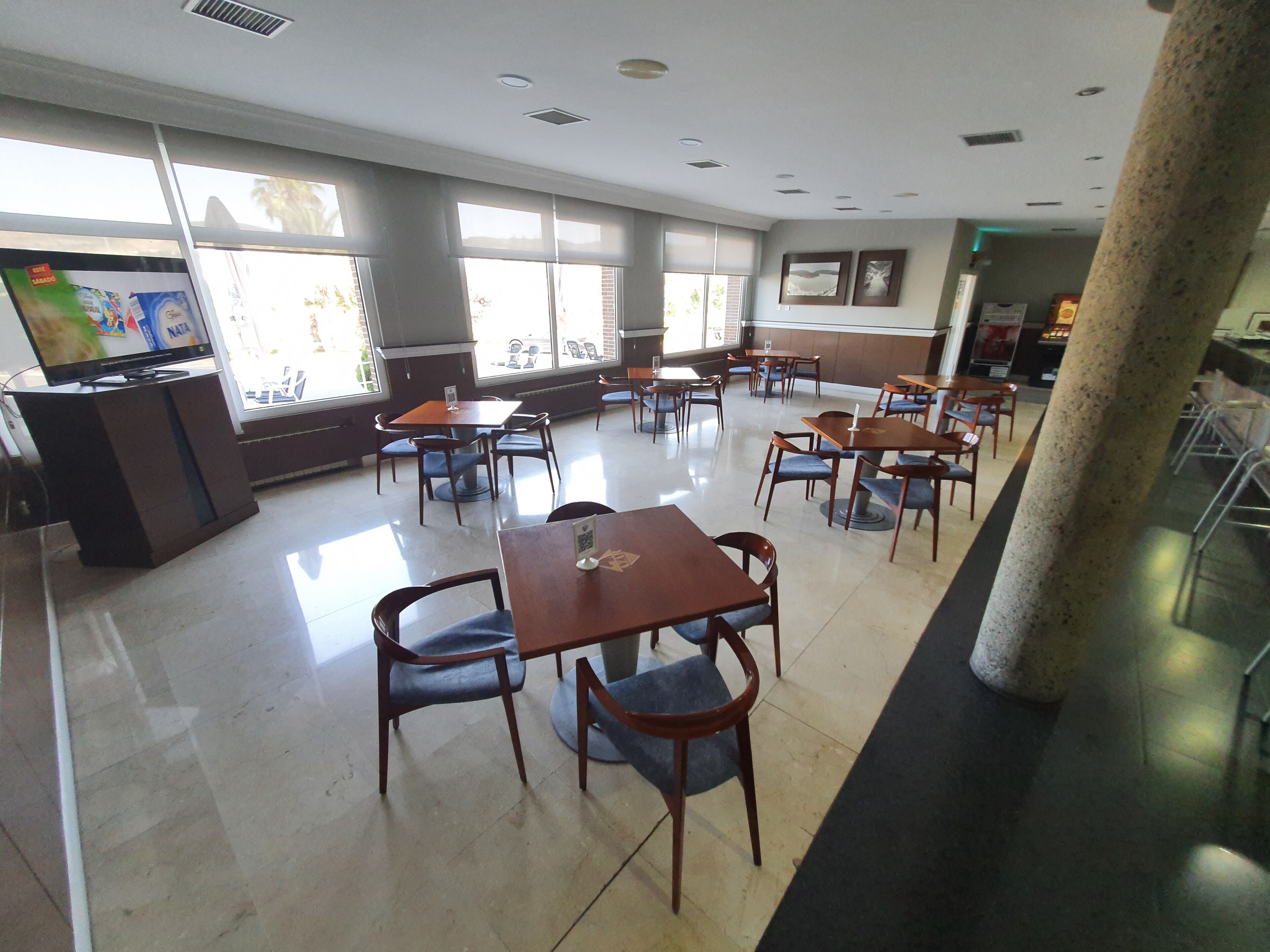 Um espaço amplo e interno com várias mesas, cadeiras, janelas grandes e um balcão com uma tela.