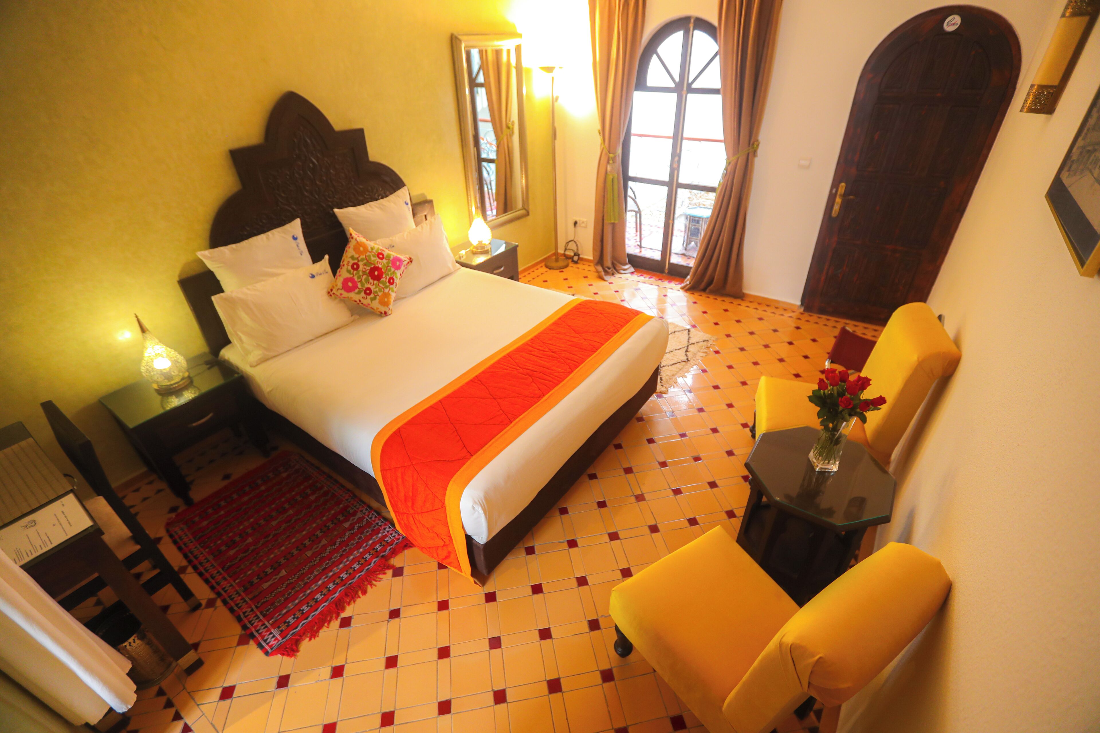 Quarto de hotel com uma cama, mesa de cabeceira, cadeira, mesa com um vaso de flores e uma janela grande com cortinas.