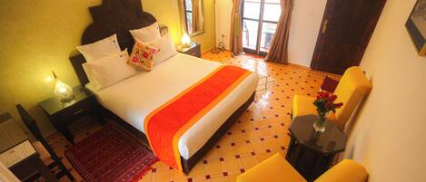 Quarto de hotel com uma cama, mesa de cabeceira, cadeira, mesa com um vaso de flores e uma janela grande com cortinas.