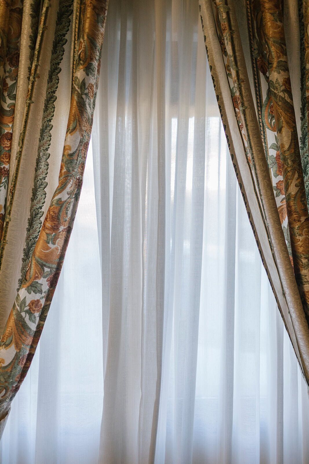 Uma janela com cortinas finas brancas e cortinas elaboradas com padrão floral.