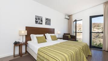 Quarto de hotel com uma cama grande, duas mesas de cabeceira, uma cadeira e uma janela ampla com vista para o deserto.