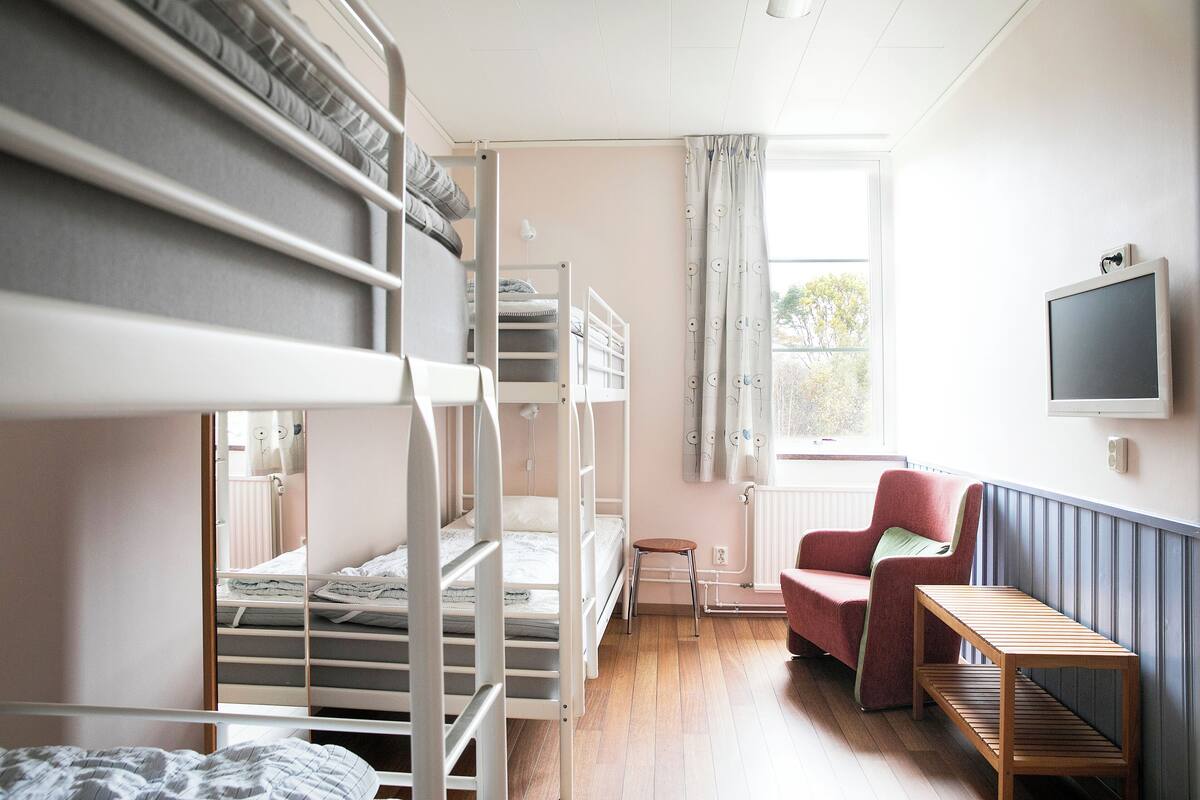 Quarto com beliche, uma pequena mesa, uma cadeira, janela com cortinas e televisão na parede.