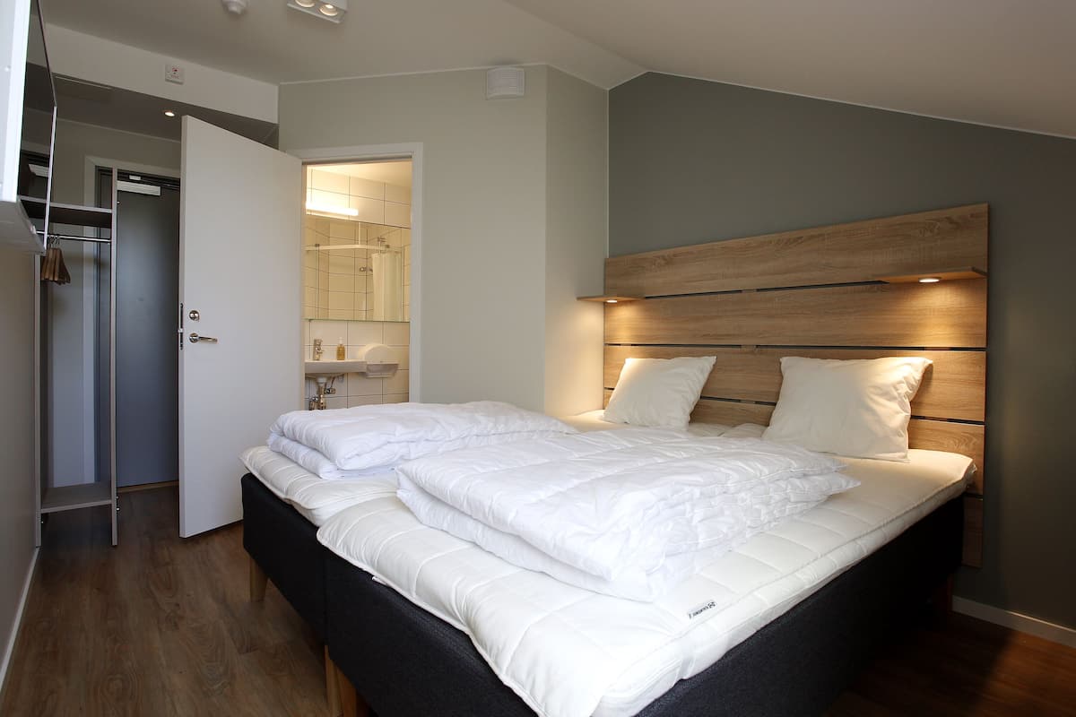 Quarto com cama, cabeceira de madeira, guarda-roupa e banheiro visível através de uma porta aberta.