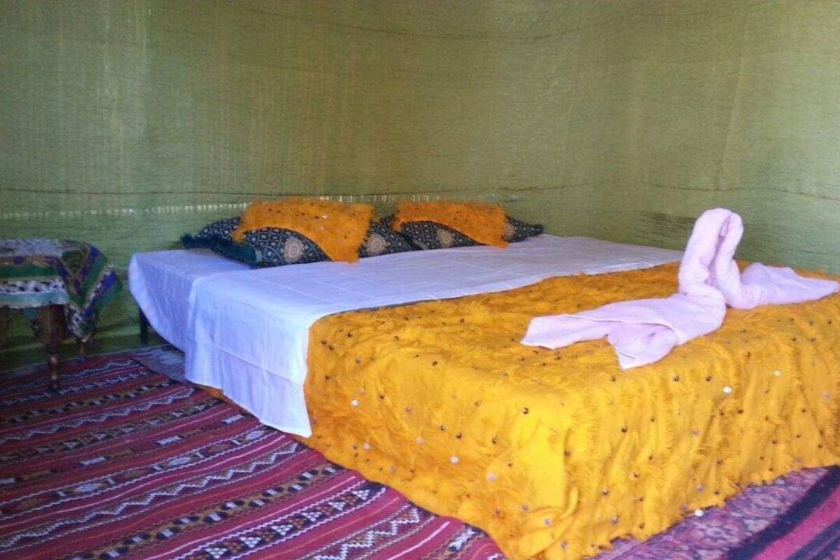 Uma cama com lençol branco, manta amarela, toalha rosa e almofadas estampadas.