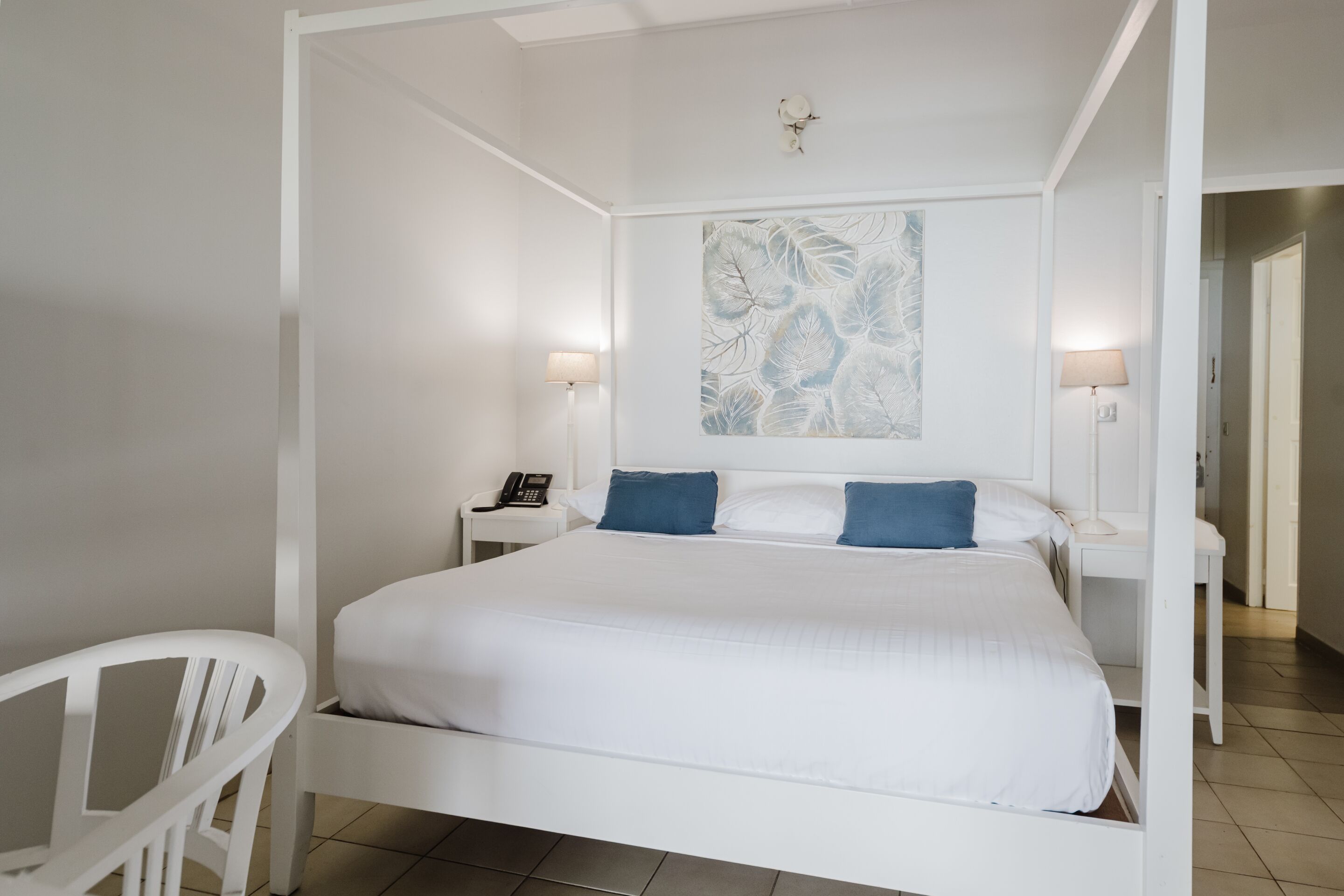 Uma cama de dossel branca com almofadas azuis, uma mesa de cabeceira com abajur e uma obra de arte pendurada na parede.