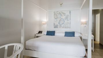 Uma cama de dossel branca com almofadas azuis, uma mesa de cabeceira com abajur e uma obra de arte pendurada na parede.