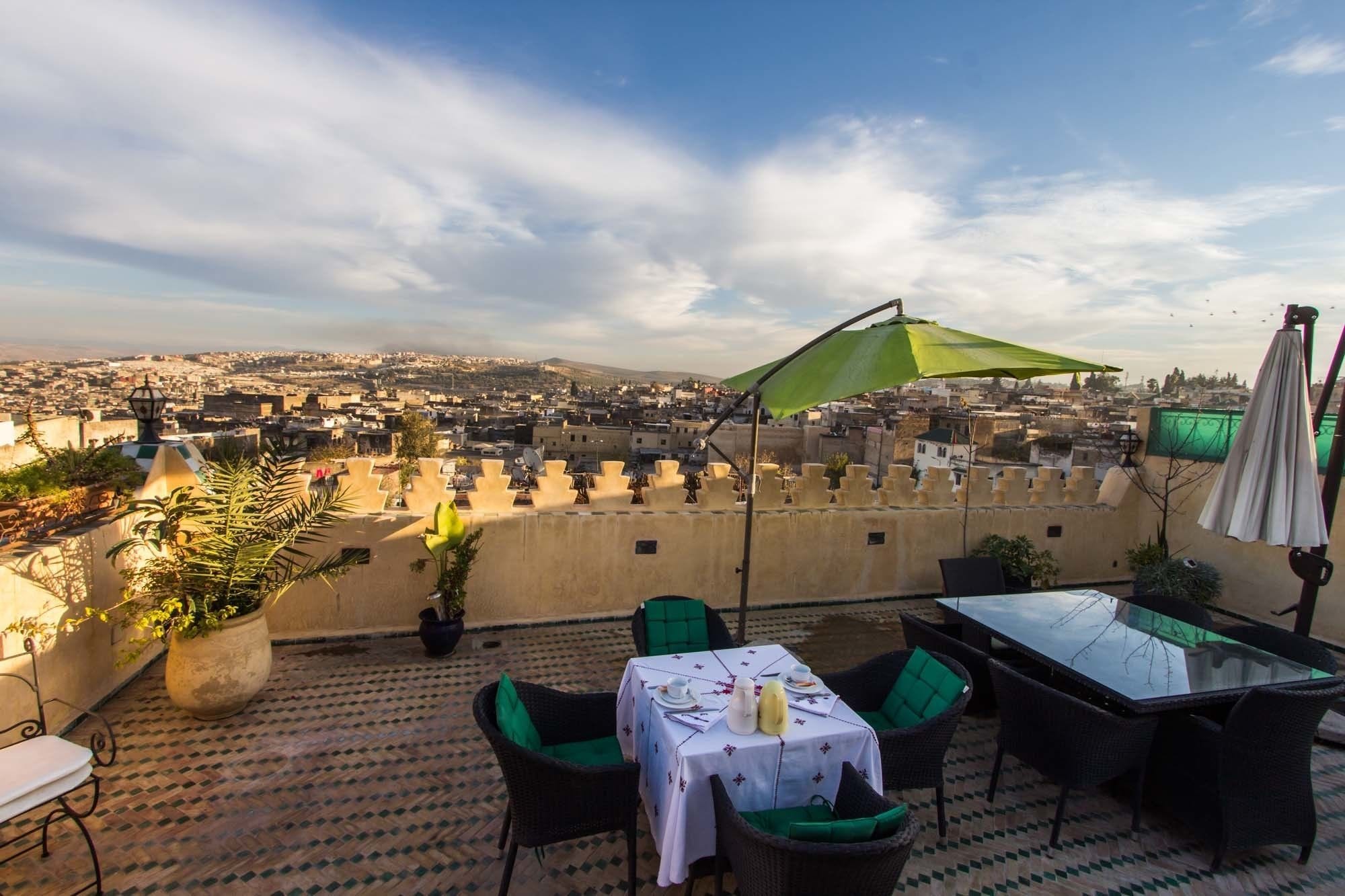 Um terraço com mesa posta para dois, um guarda-sol verde e vista para a cidade.
