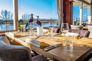 Mesa de jantar posta com copos de vinho, talheres e um menu, com vista para um lago e aves.