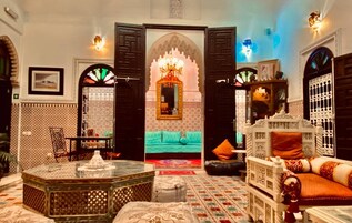 Um interior tradicional marroquino com mobiliário ornamentado, um arco central e azulejos estampados.