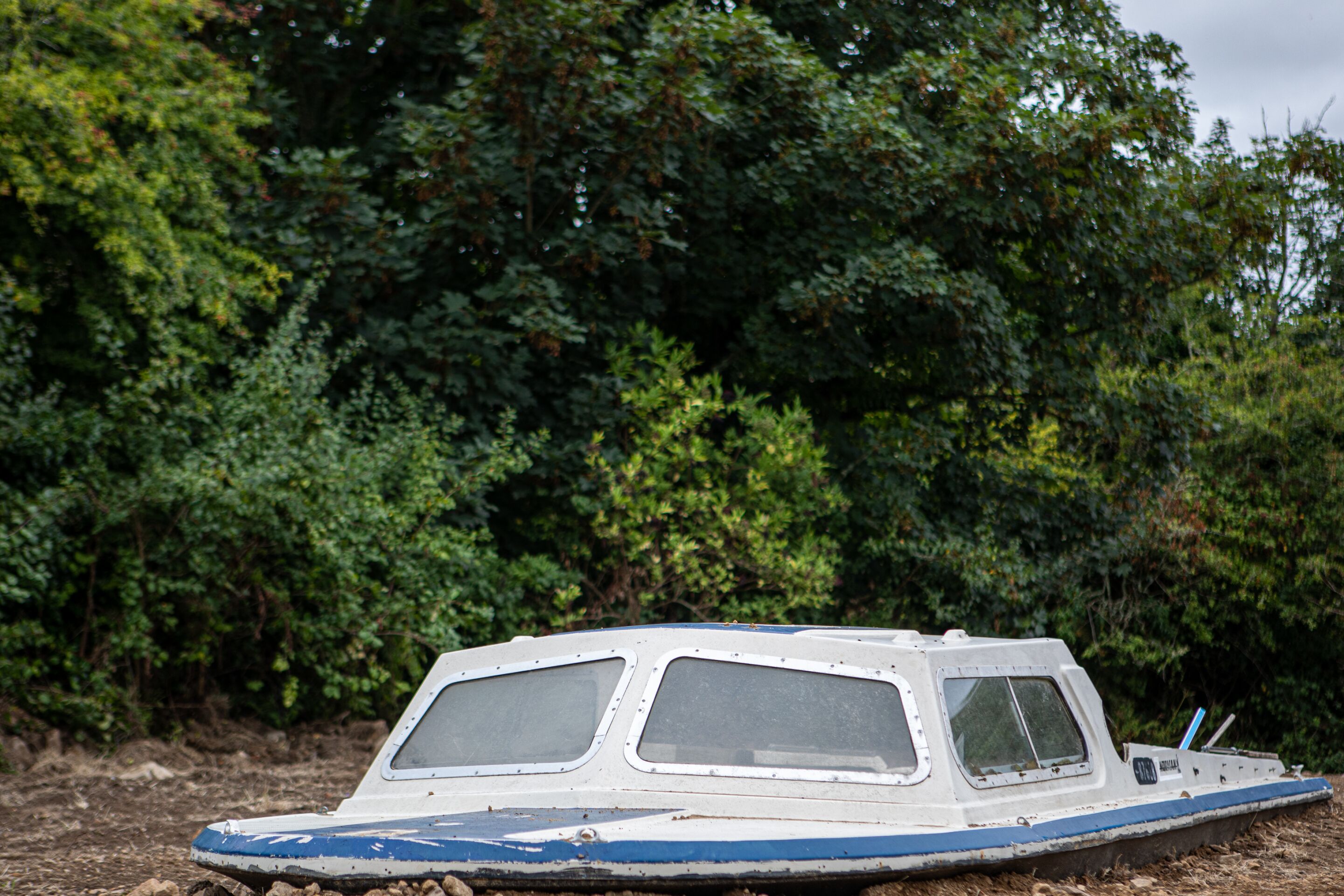 Um barco com estrutura de cabine, posicionado sobre terreno de terra batida com vegetação verde densa ao fundo.