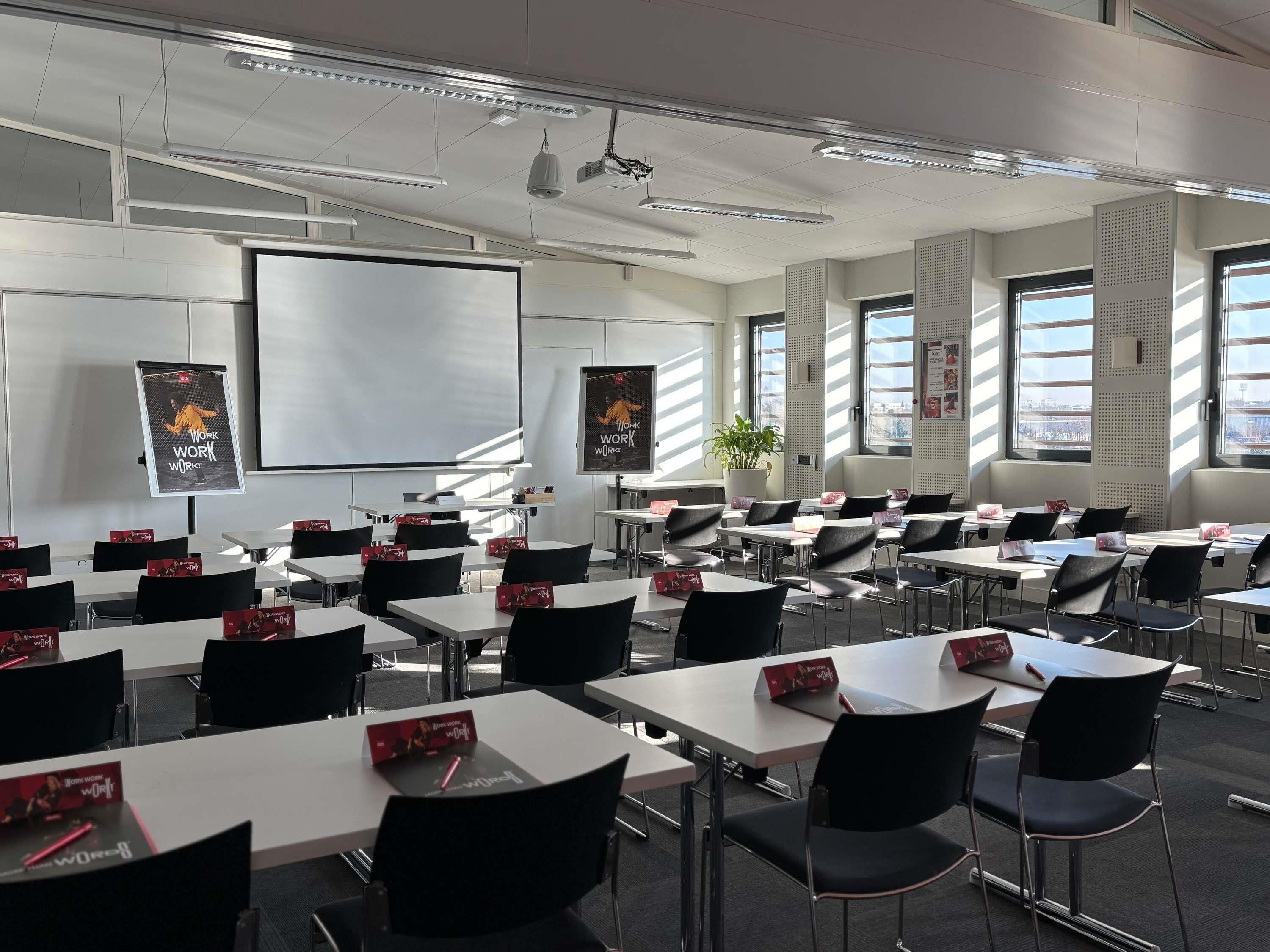 Uma sala de conferências com mesas, cadeiras, tela de projetor e cartazes nas paredes.
