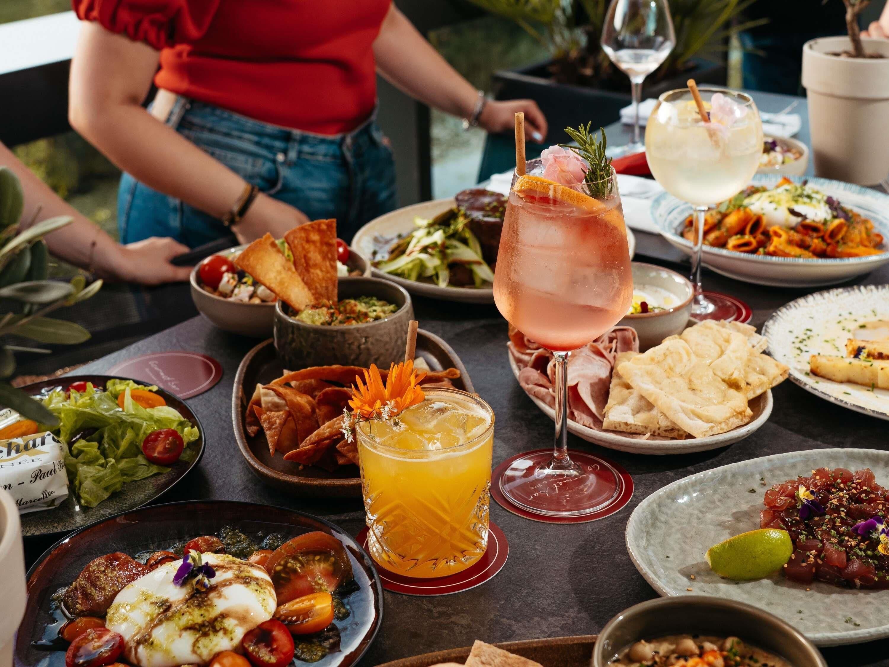 Uma mesa com diversos pratos e bebidas, incluindo uma salada, carne grelhada e um cocktail com enfeite de rosa.
