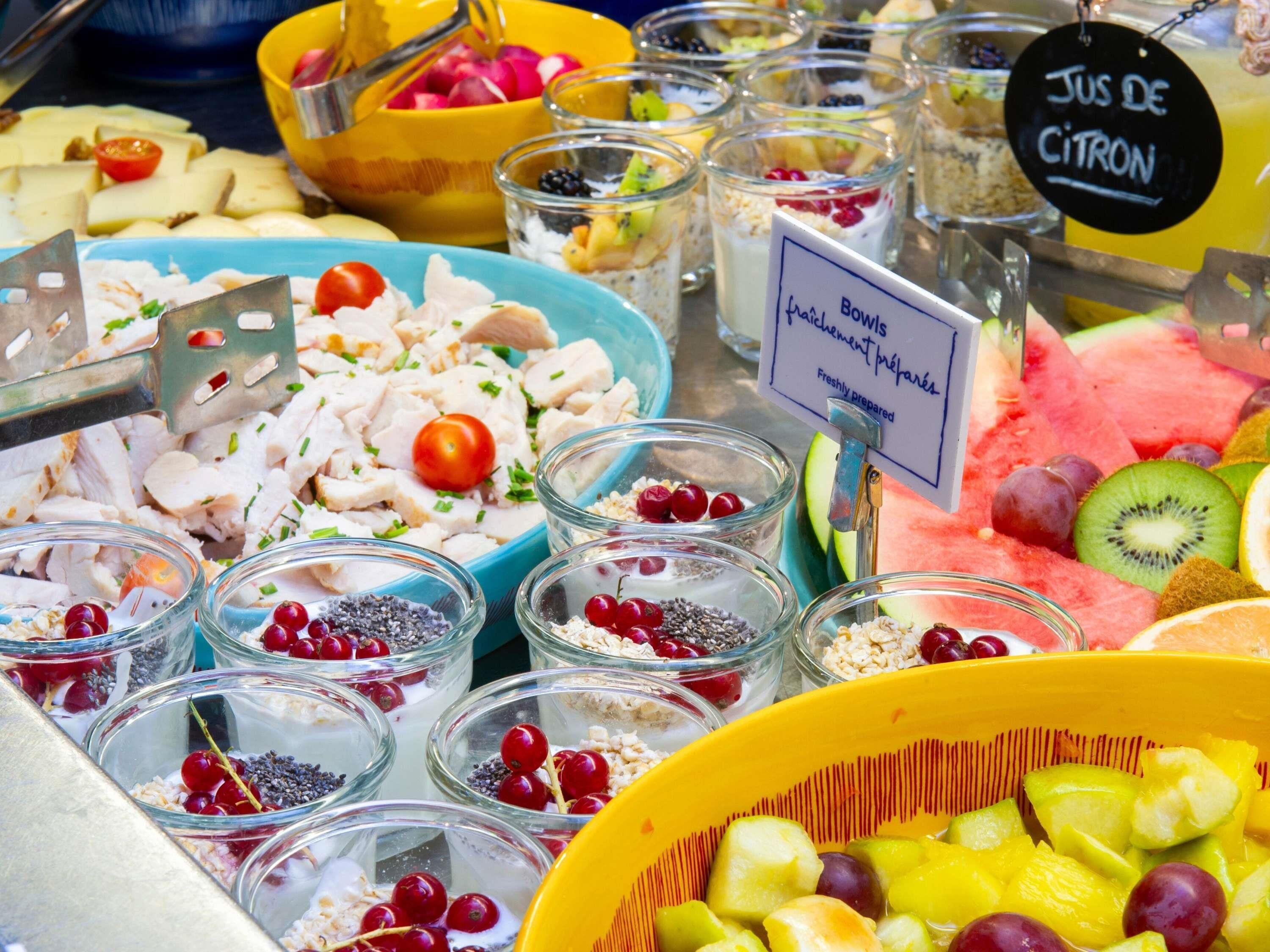 Diversos alimentos, incluindo saladas em tigelas, frutas e um aviso indicando pratos 'preparados na hora'.