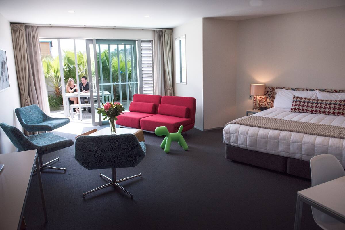 Quarto de hotel com uma cama, um sofá vermelho, uma poltrona azul, um brinquedo de cachorro verde e uma varanda com vista para palmeiras.