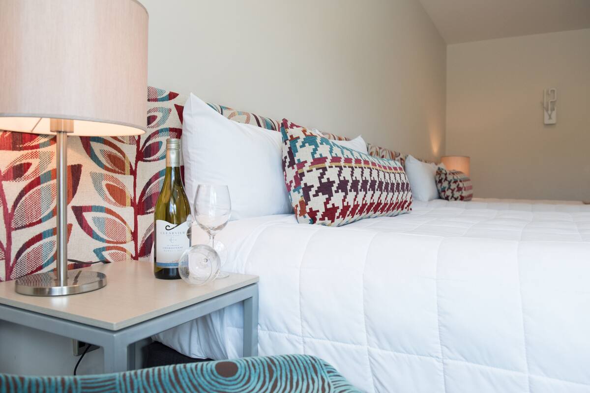 Quarto de hotel bem arrumado com cama, almofadas, mesa de cabeceira com luminária, garrafa de vinho e um copo.