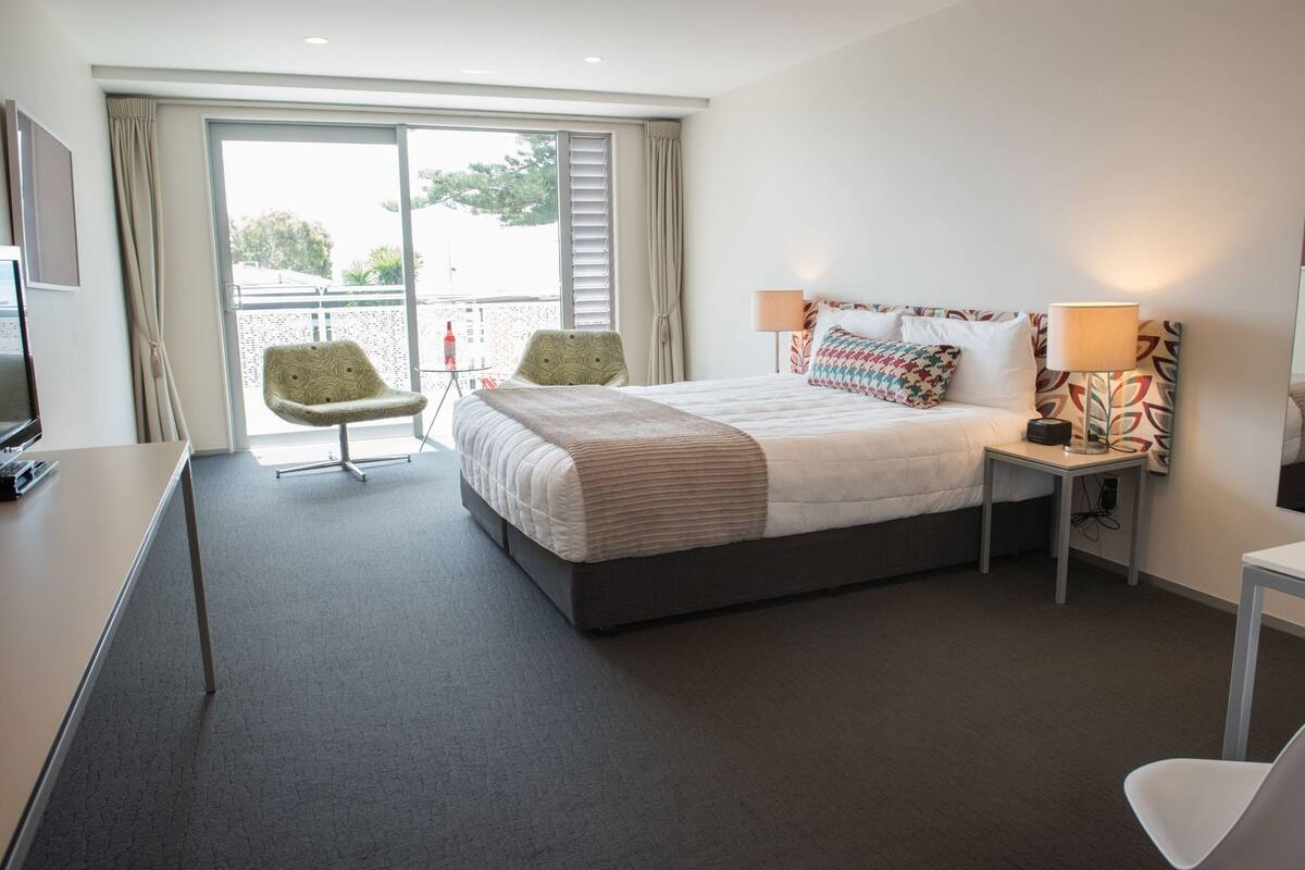 Quarto de hotel com uma cama, uma escrivaninha, duas cadeiras, uma televisão e varanda com cortinas.