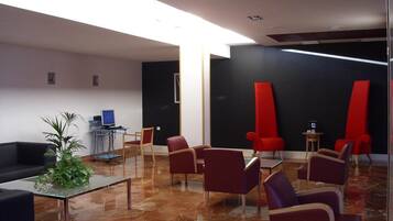 Um lobby moderno com uma mesa de centro de vidro, cadeiras vermelhas e pretas, uma mesa de escritório com um computador e uma planta em vaso.